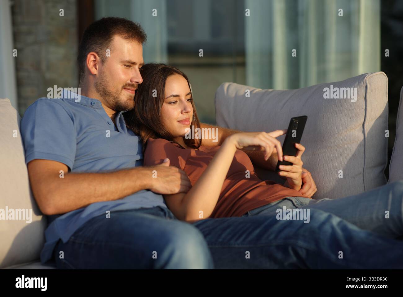 Relaxed couple at sunset checking mobile phone sitting on a couch in a ...