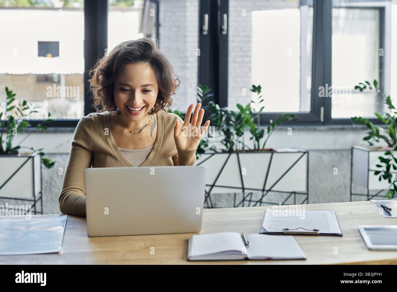 A person cheerfully waves at colleagues while using a laptop in a ...