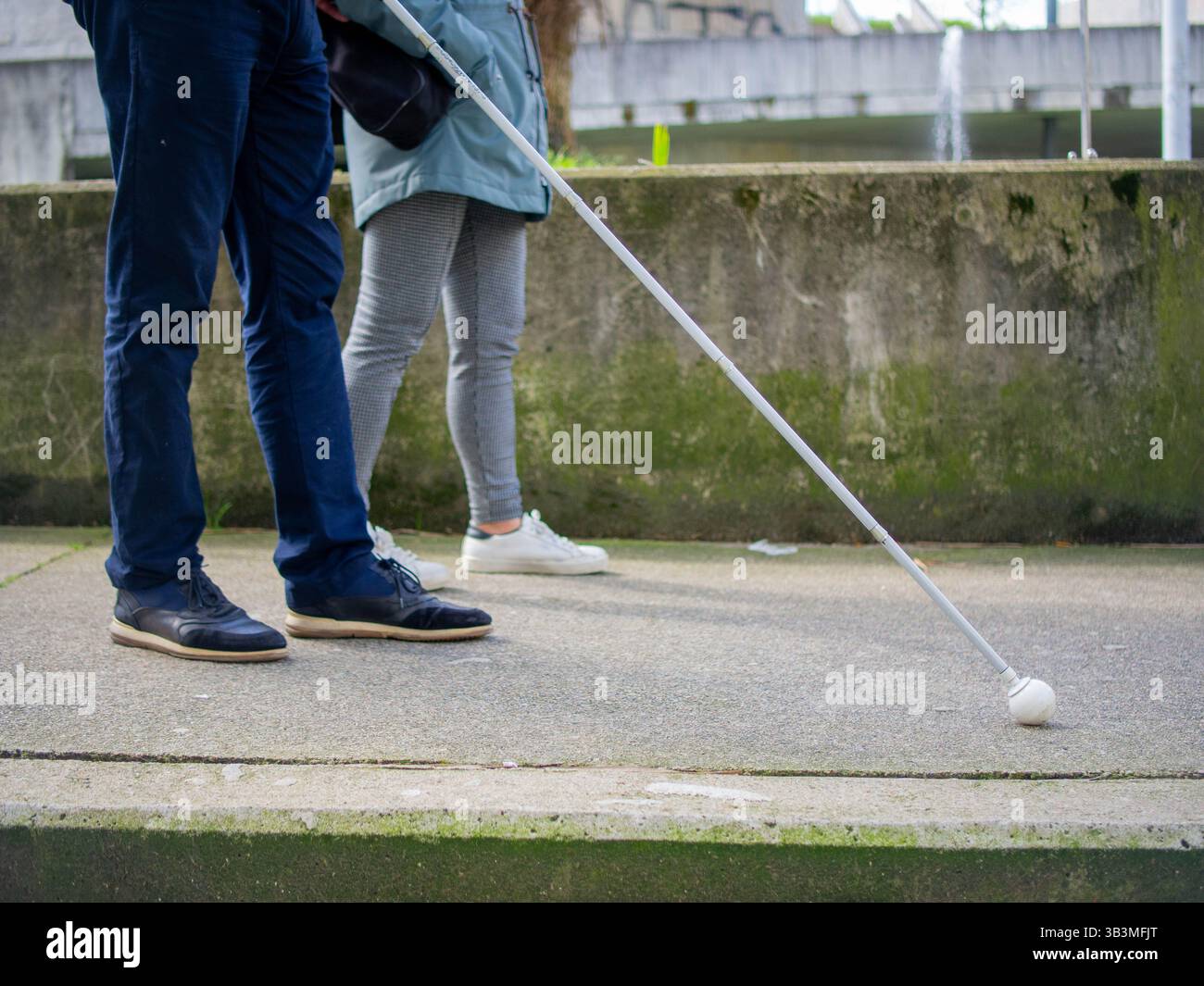 Blind man walking on the pavement using his cane and a woman providing ...