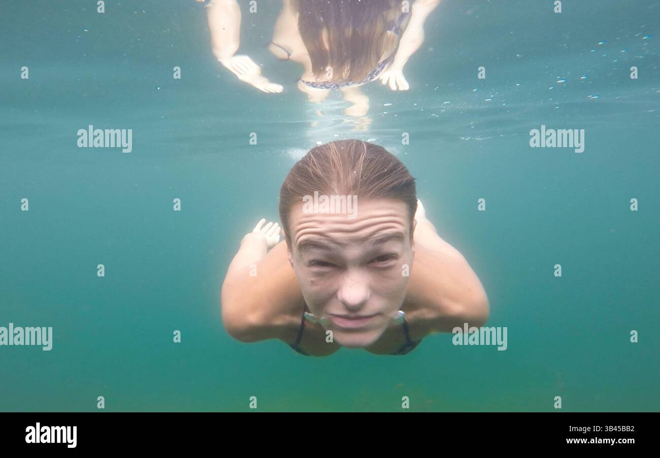 Young woman is swimming under the water towards the camera looking into ...