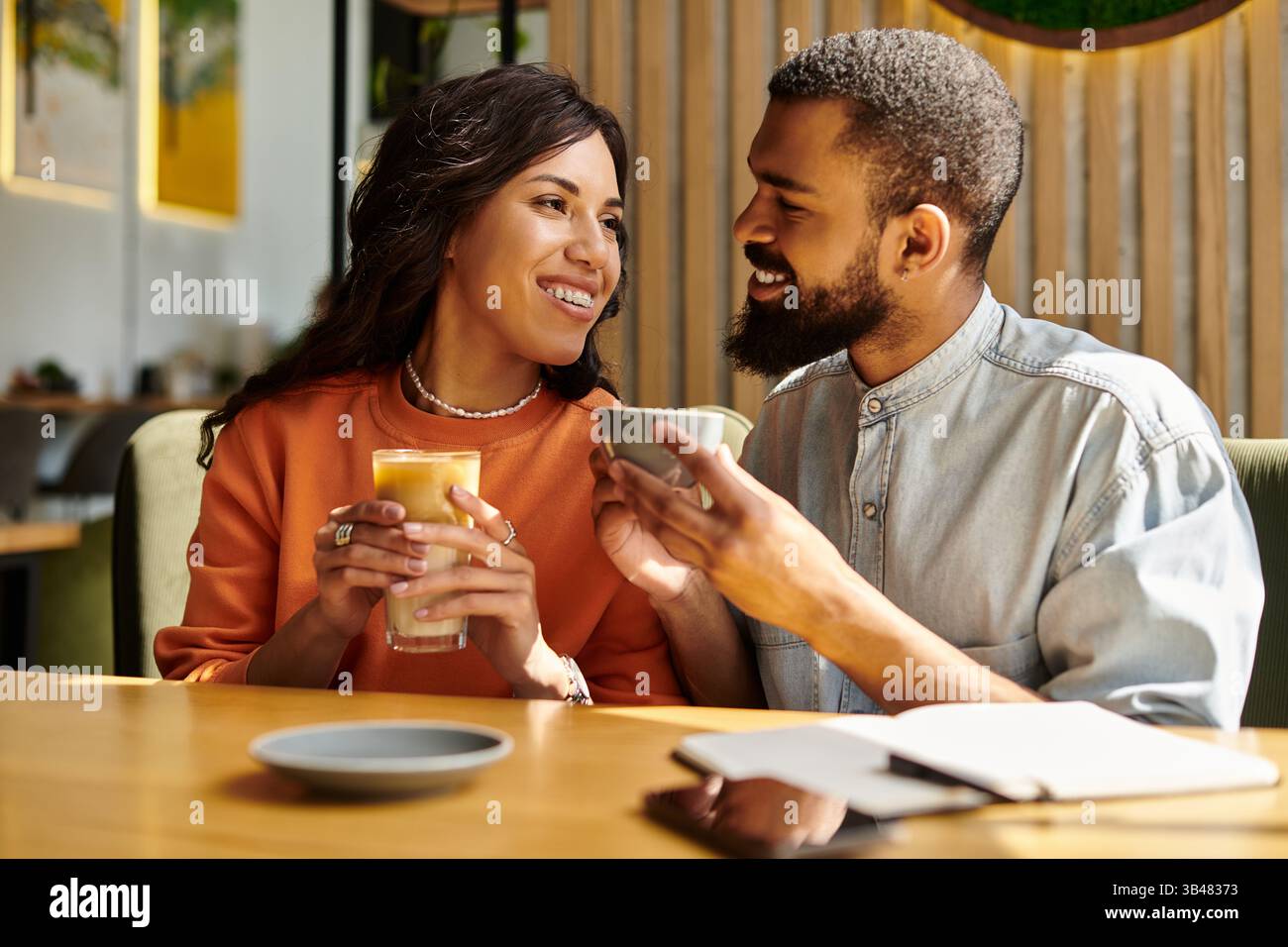 An African American couple shares laughter and drinks in a warm cafe setting, radiating joy ...