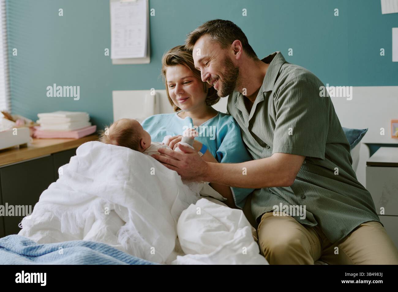Smiling parents holding their newborn baby while sitting on bed in ...