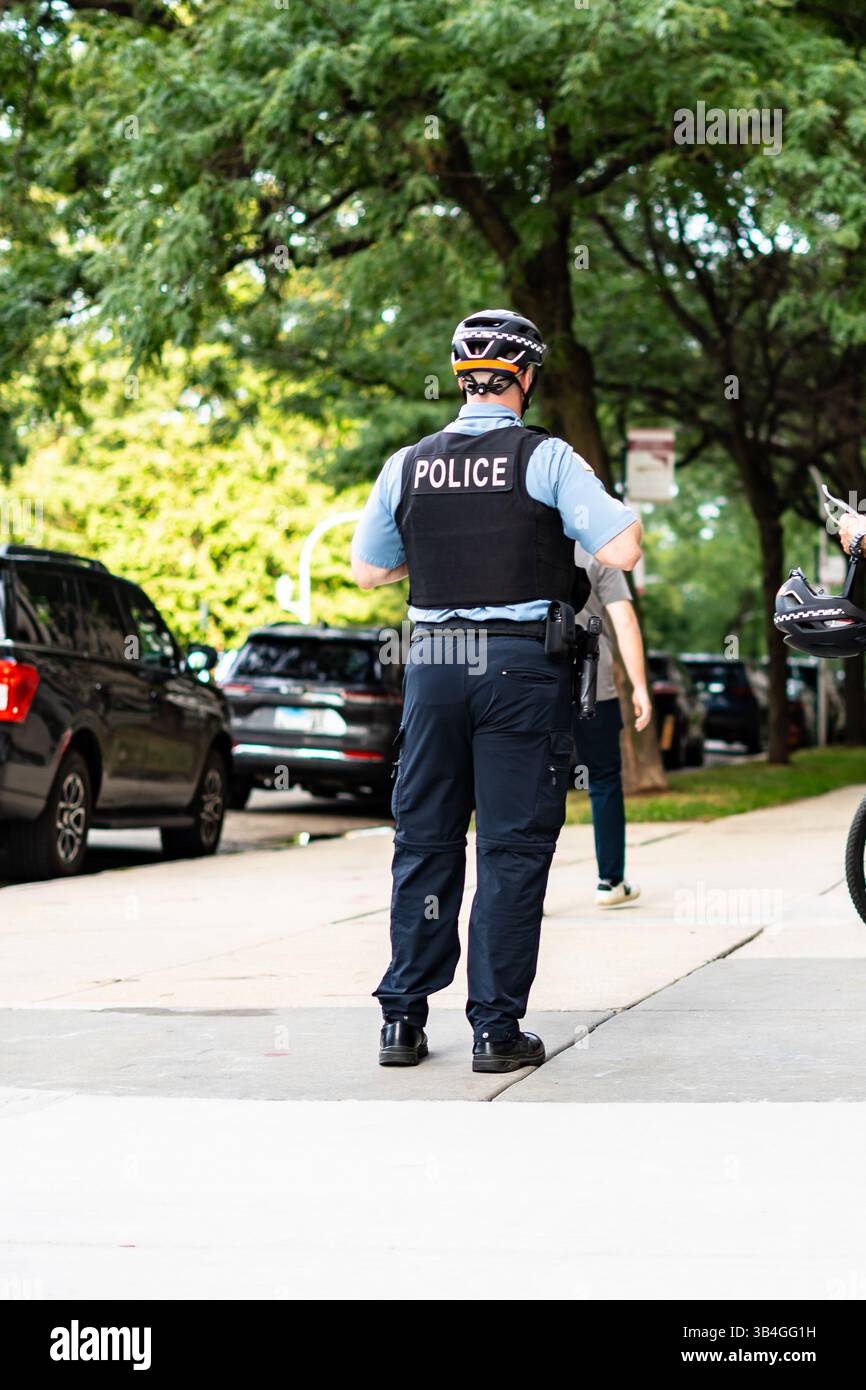 Uniformed police officer on patrol in an urban neighborhood, standing near a city sidewalk ...