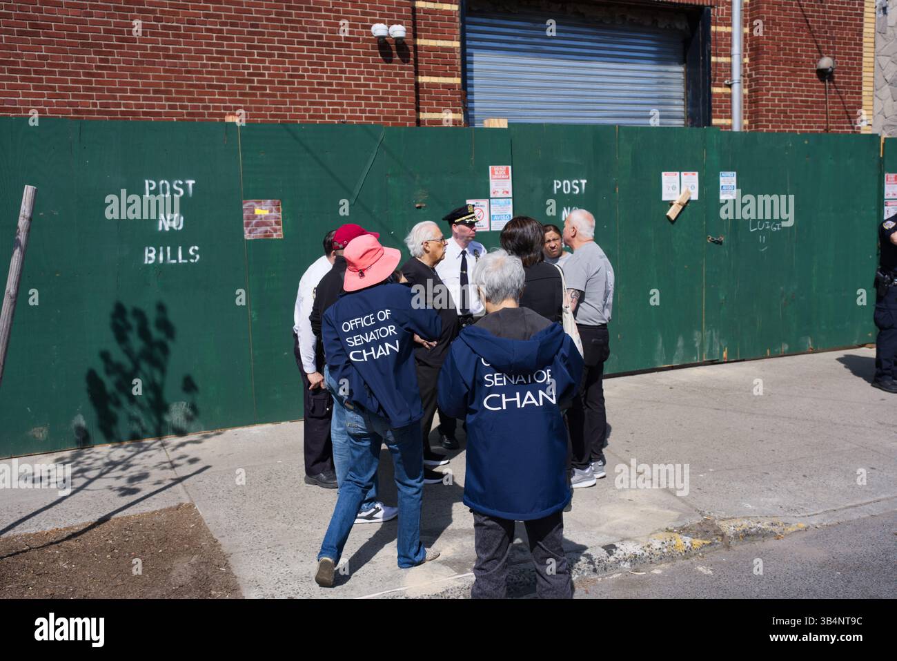 A group of people stand before a green construction fence. Several wear ...