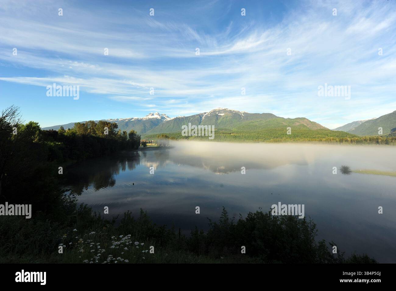Morning mist drifts over a still mountain lake, reflecting blue skies ...