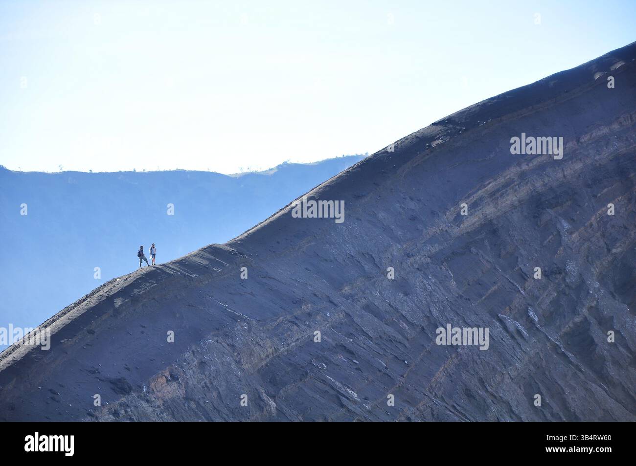 Two hikers walk along the rim of a volcanic crater with dramatic slopes ...