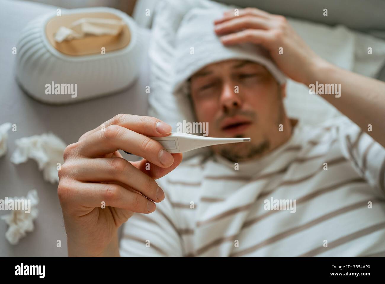 Sick man suffering from cold or flu, lying in bed with wet towel on his ...