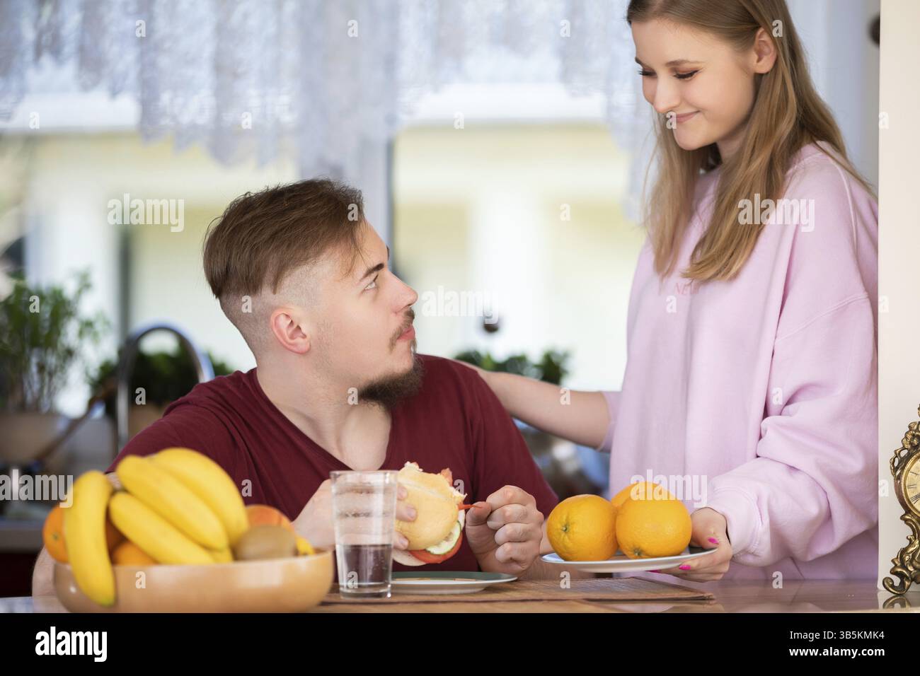 The boy is eating breakfast in the kitchen. A smiling blonde girl enters the room and brings a ...