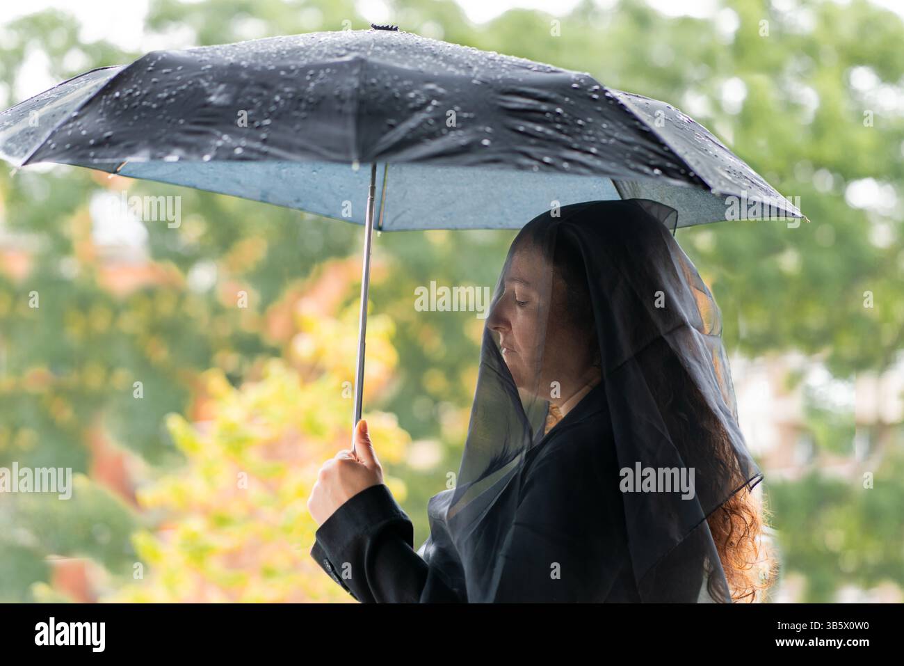 Side view of sad woman in black veil with umbrella in cemetery for ...