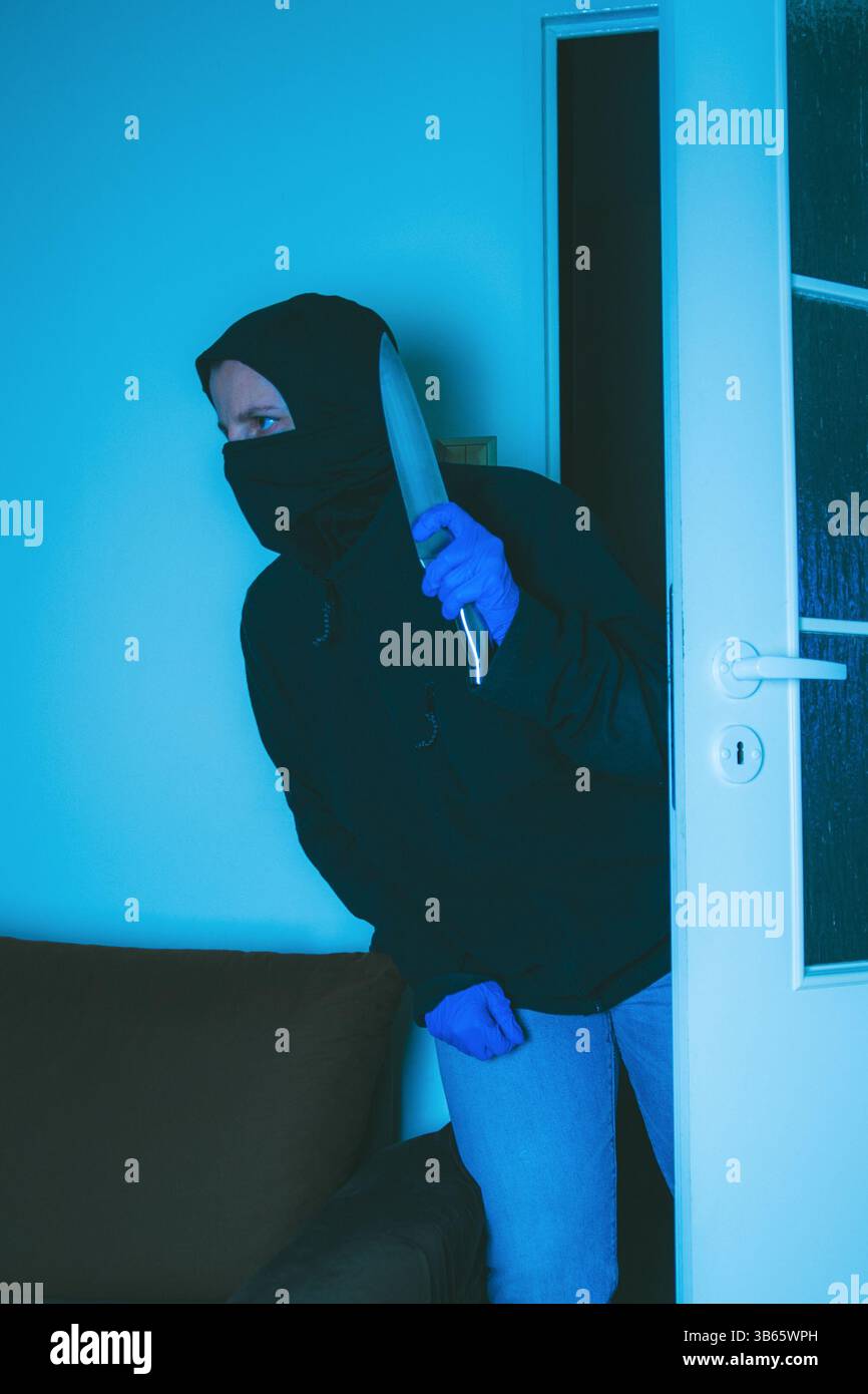 Masked intruder wearing black clothing, holding a knife, peeking ...