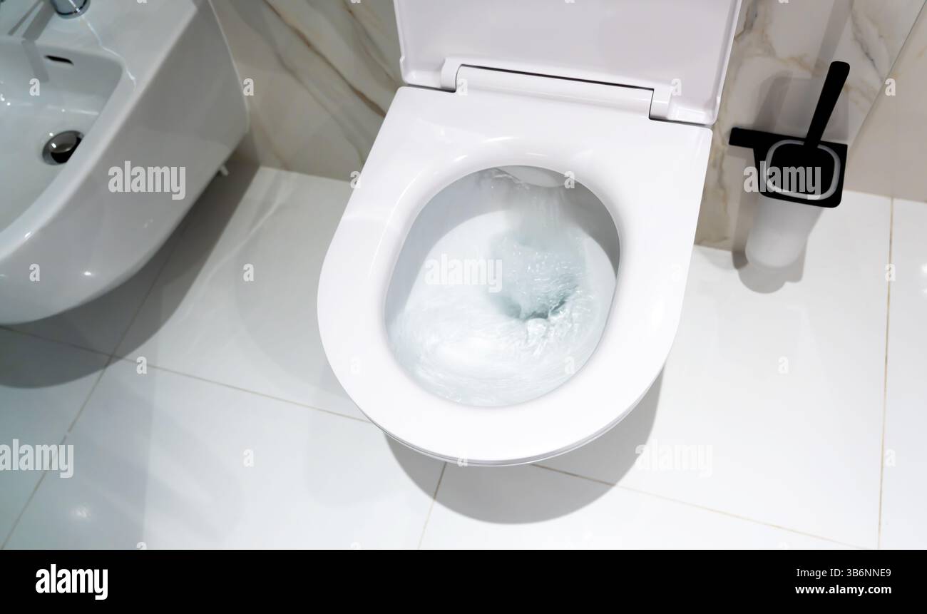 A porcelain toilet flushing with clean swirling water in a tidy white ...