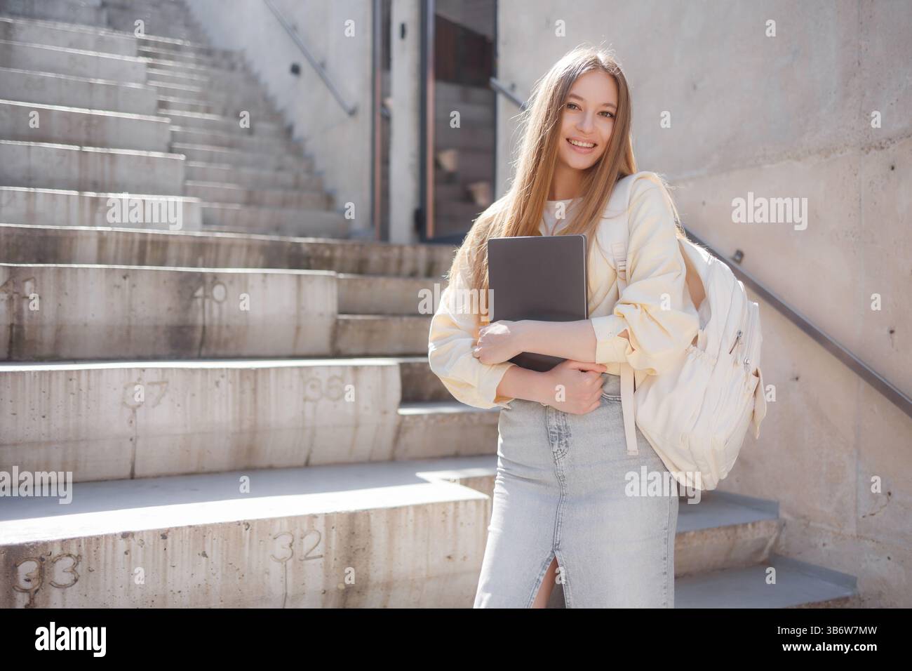 Student poses on outdoor stairs, smiling while holding laptop ...