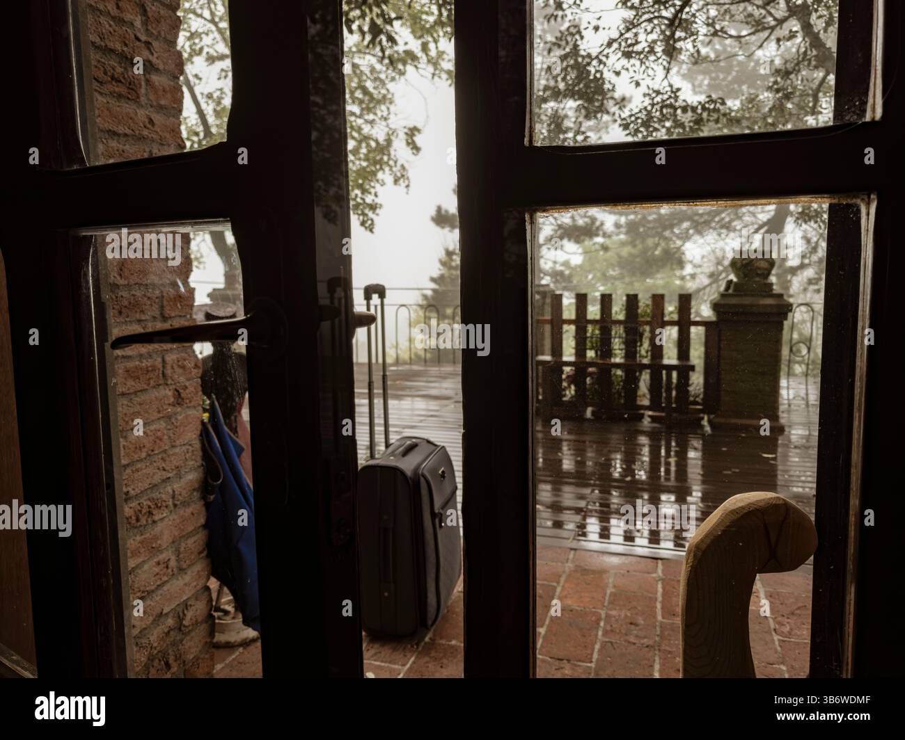 Black suitcase and blue umbrella waiting inside by the door while rain is pouring outside on a ...
