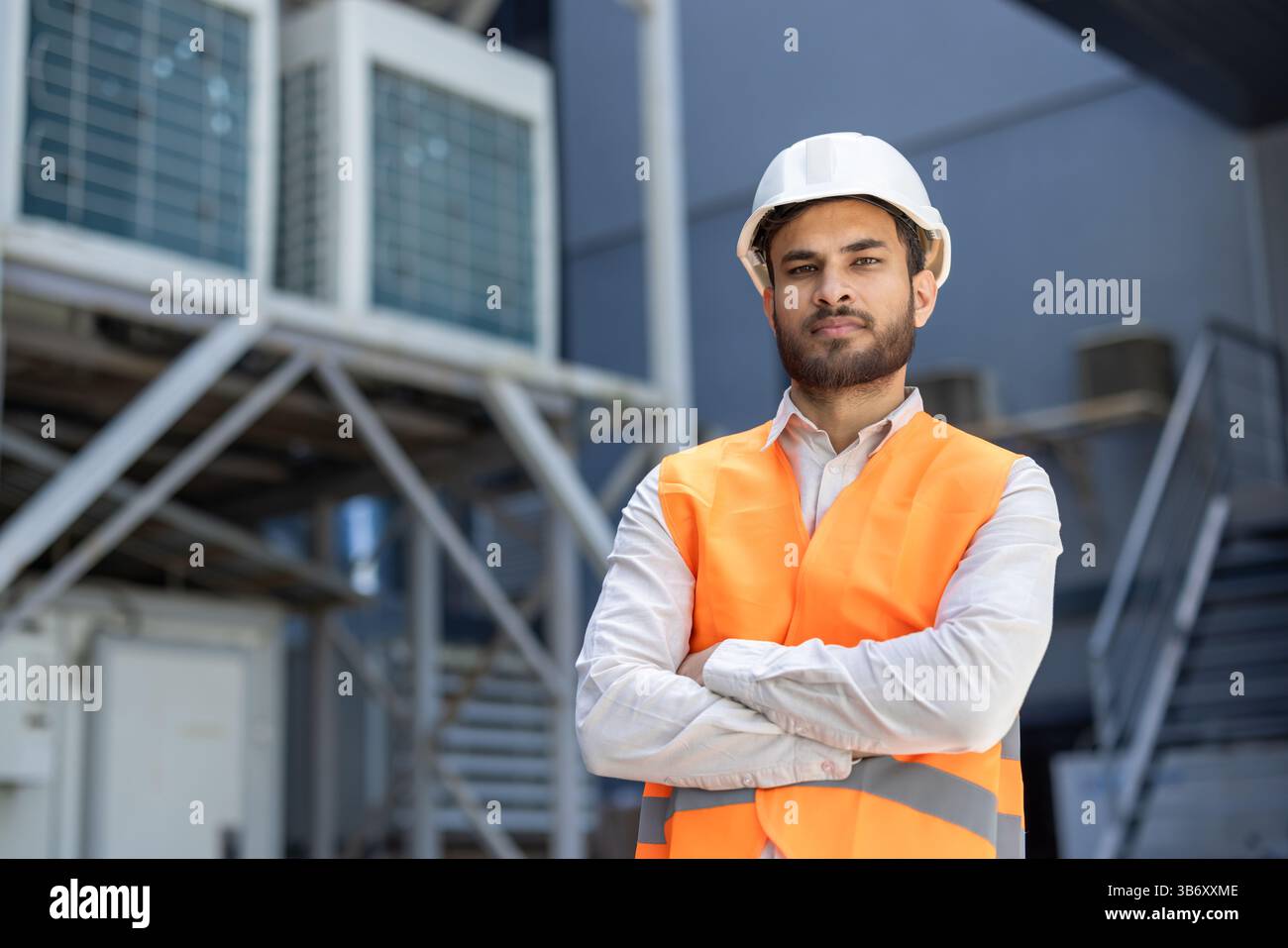 A construction worker in a hard hat and safety vest stands confidently outdoors, arms crossed ...