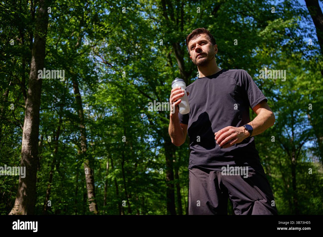 Man wearing black sportswear drinking water during run in forest ...
