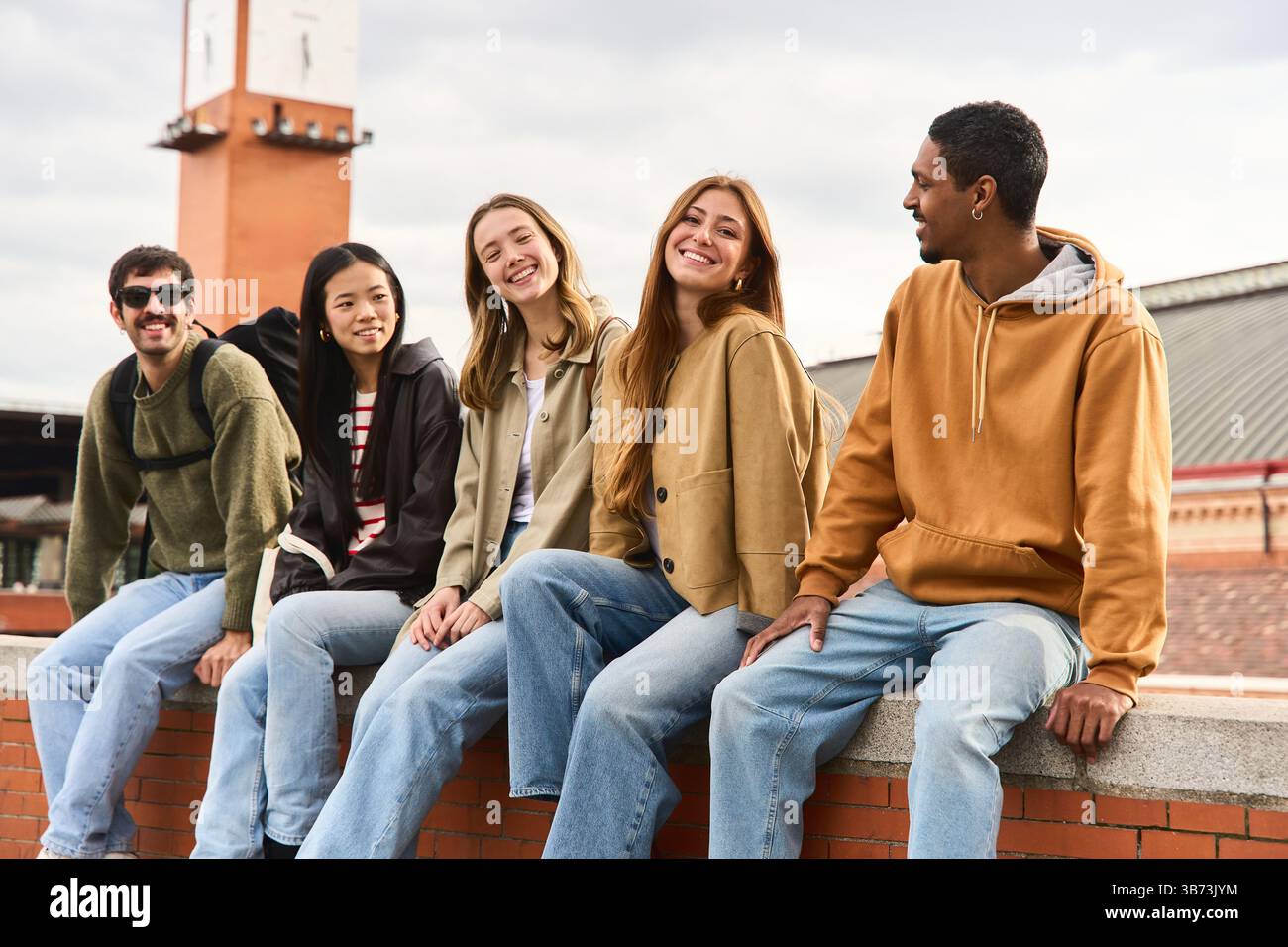 Five happy young students are sitting on a brick wall, talking and ...