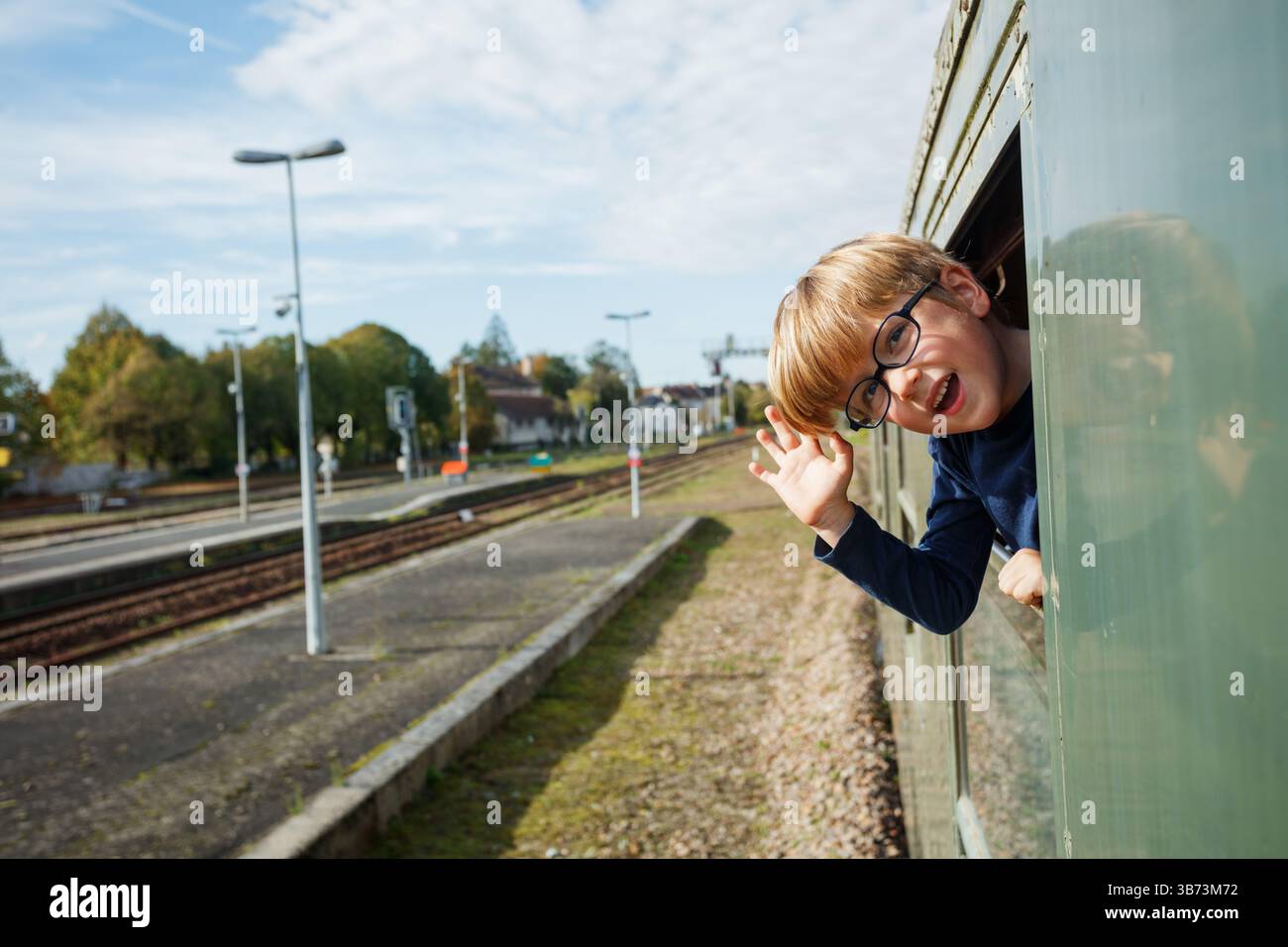 A blond boy in blue leans from a green carriage window, waving ...