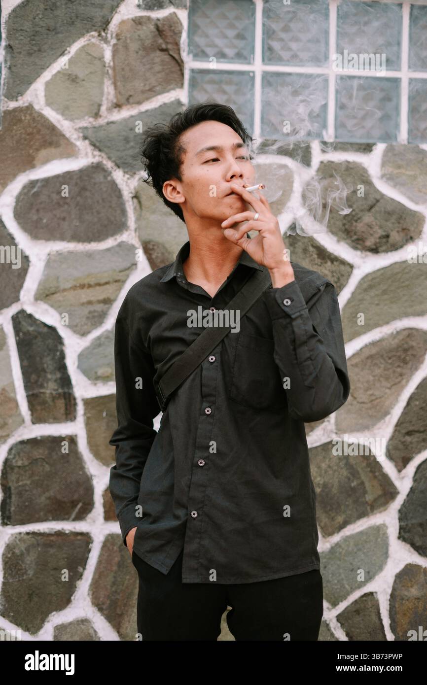Stylish asian man smoking a cigarette against a stone wall. He is ...