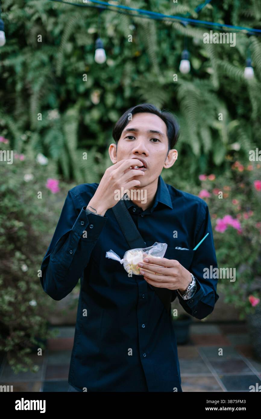 Asian man enjoying a snack outdoors with a beautiful foliage backdrop ...