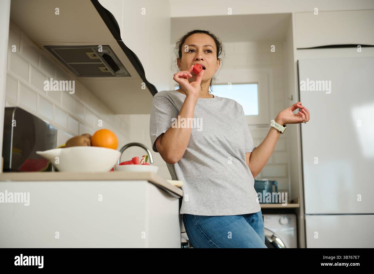 A woman enjoys eating a slice of fruit in a bright, modern kitchen ...
