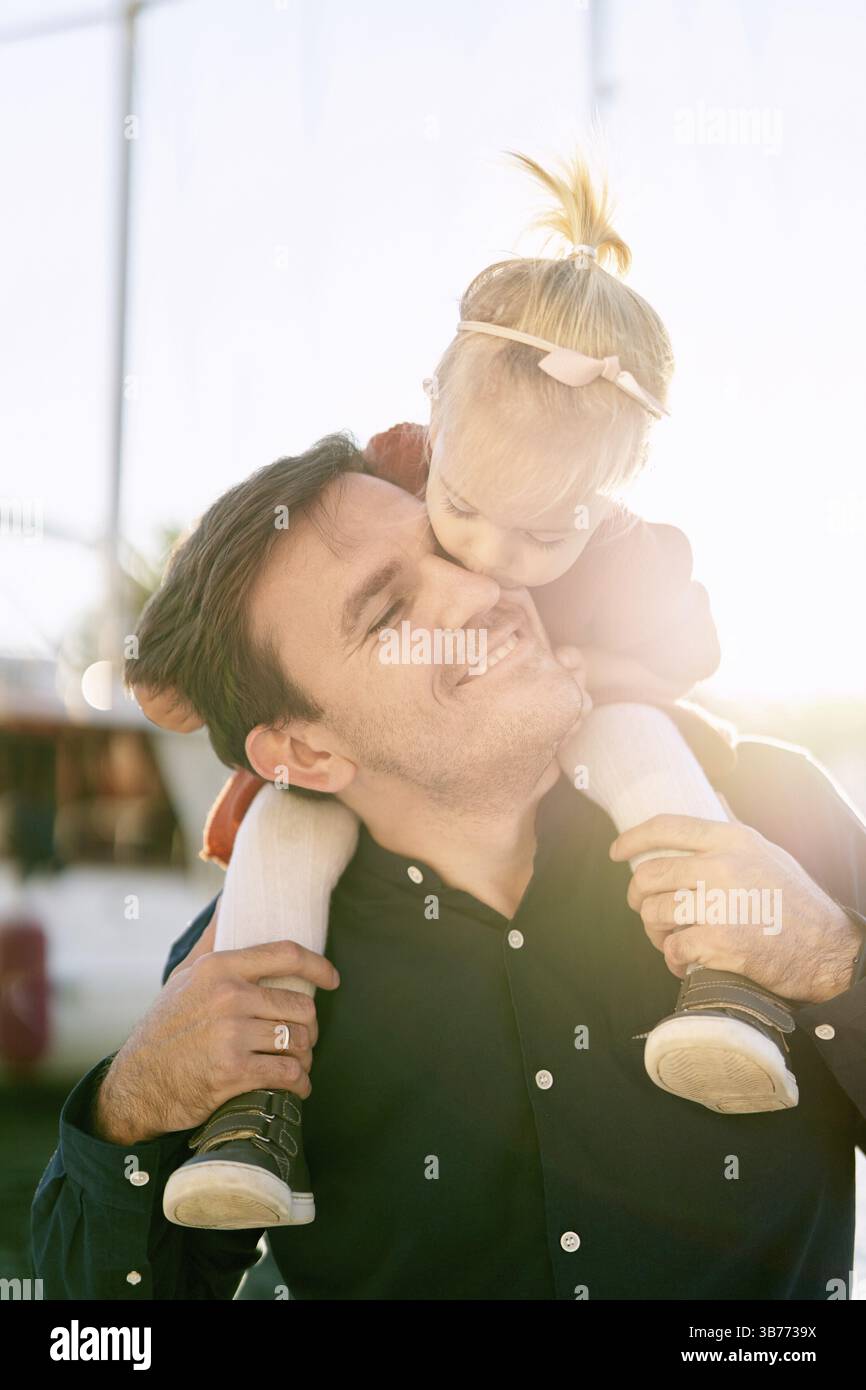 2-year old is sitting on her father's shoulders and kissing him on the ...