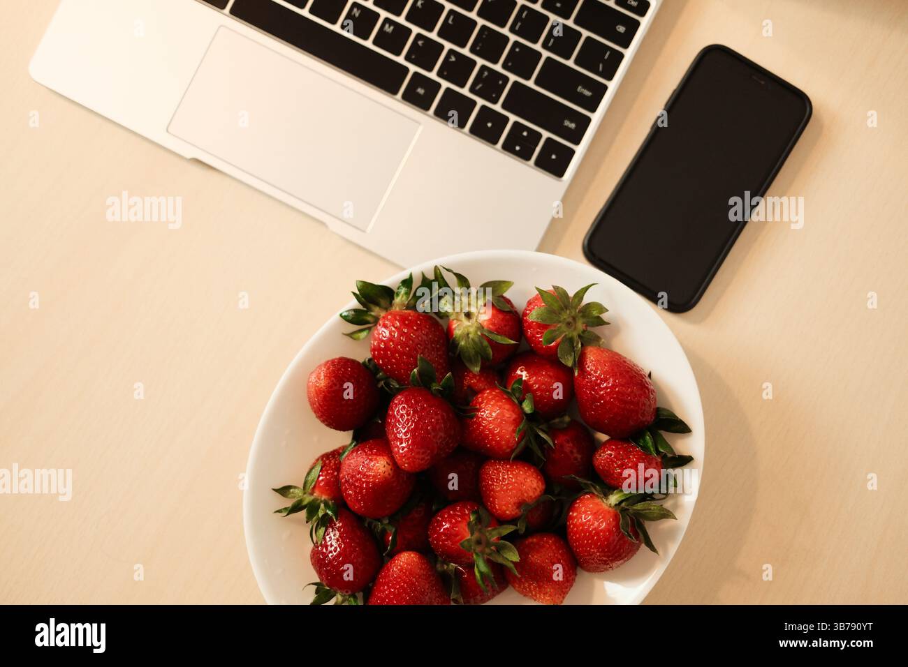 A white bowl of glossy red strawberries sits beside a sleek smartphone ...