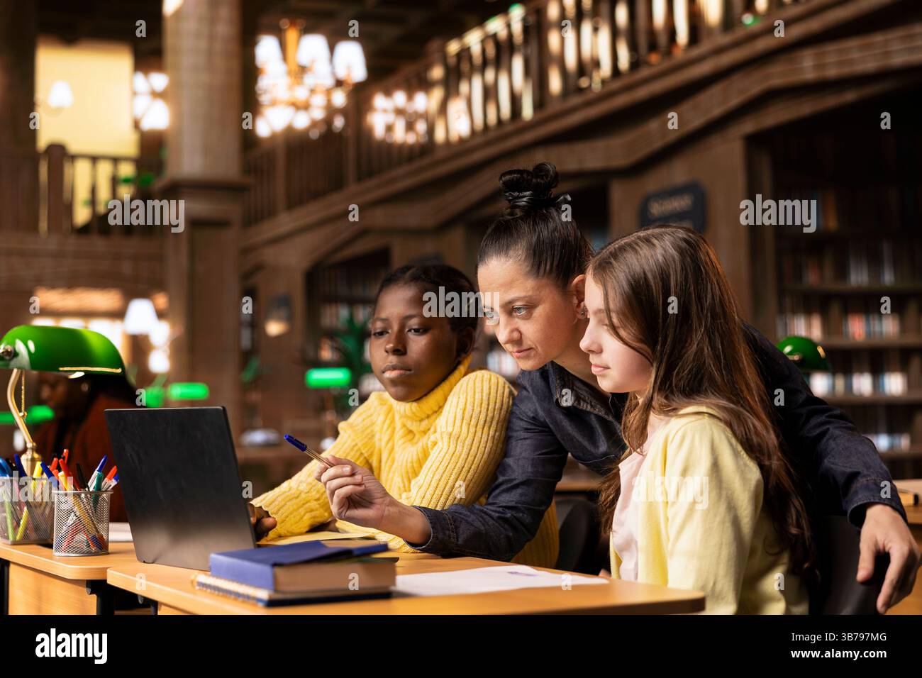 Caucasian tutor guiding two elementary school girls at a library desk ...
