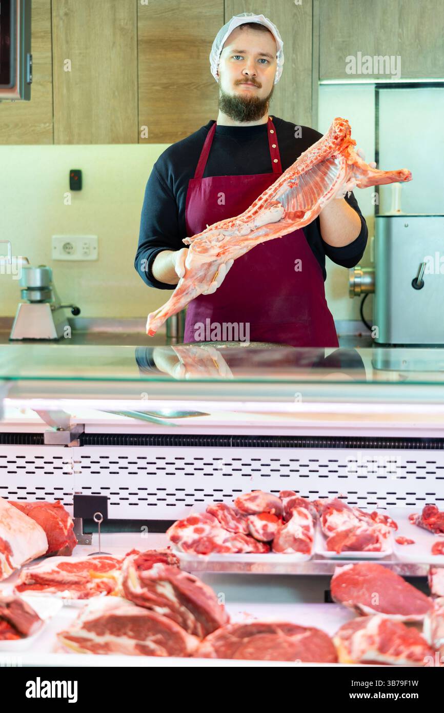 Bearded butcher demonstrating half of fresh lamb carcass at counter ...