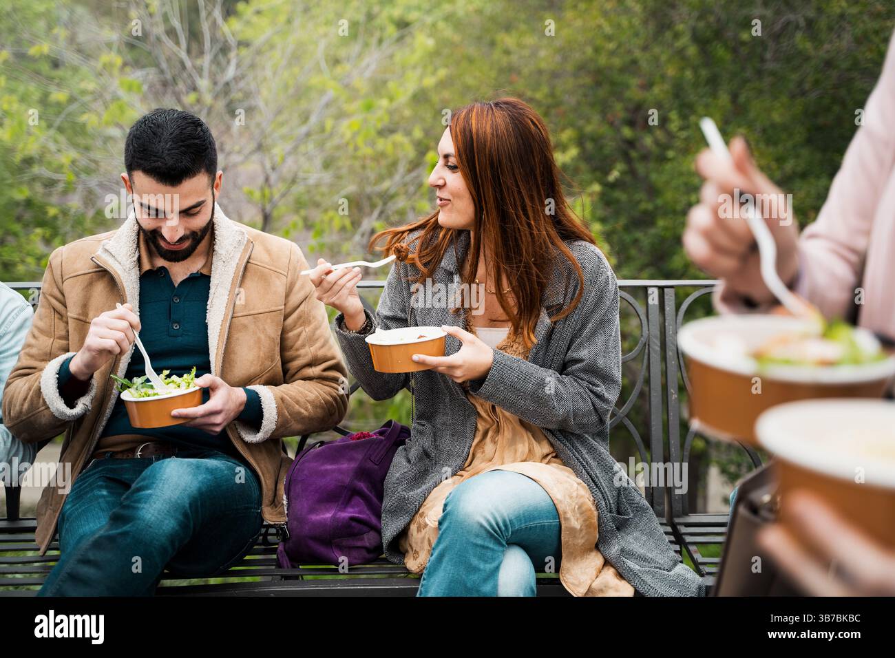 Young couple having lunch outdoors on a park bench, sharing a relaxed ...