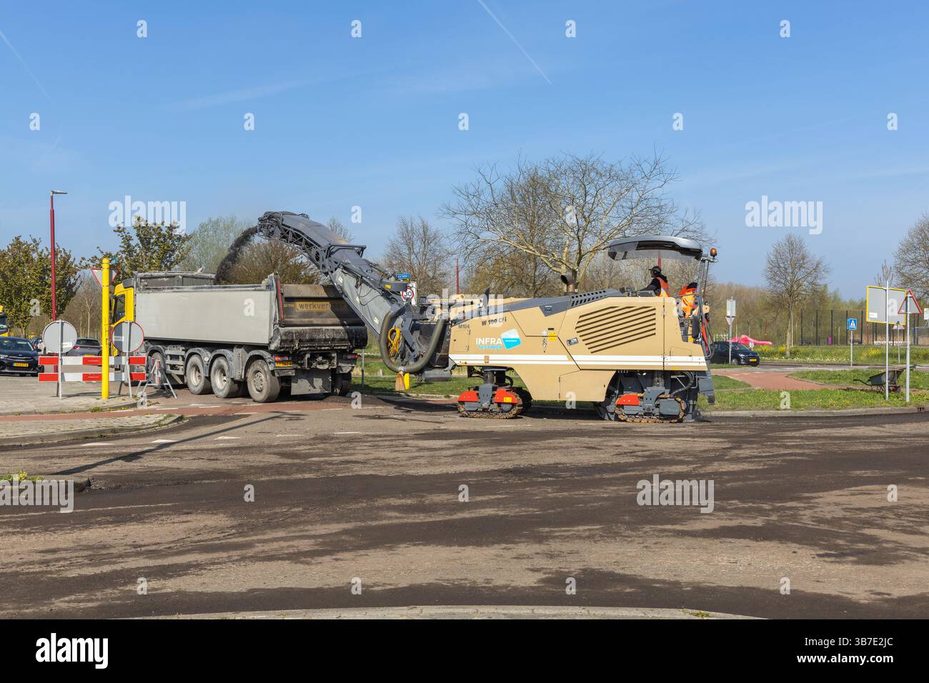 A milling machine removes asphalt from a road, loading it into a truck ...