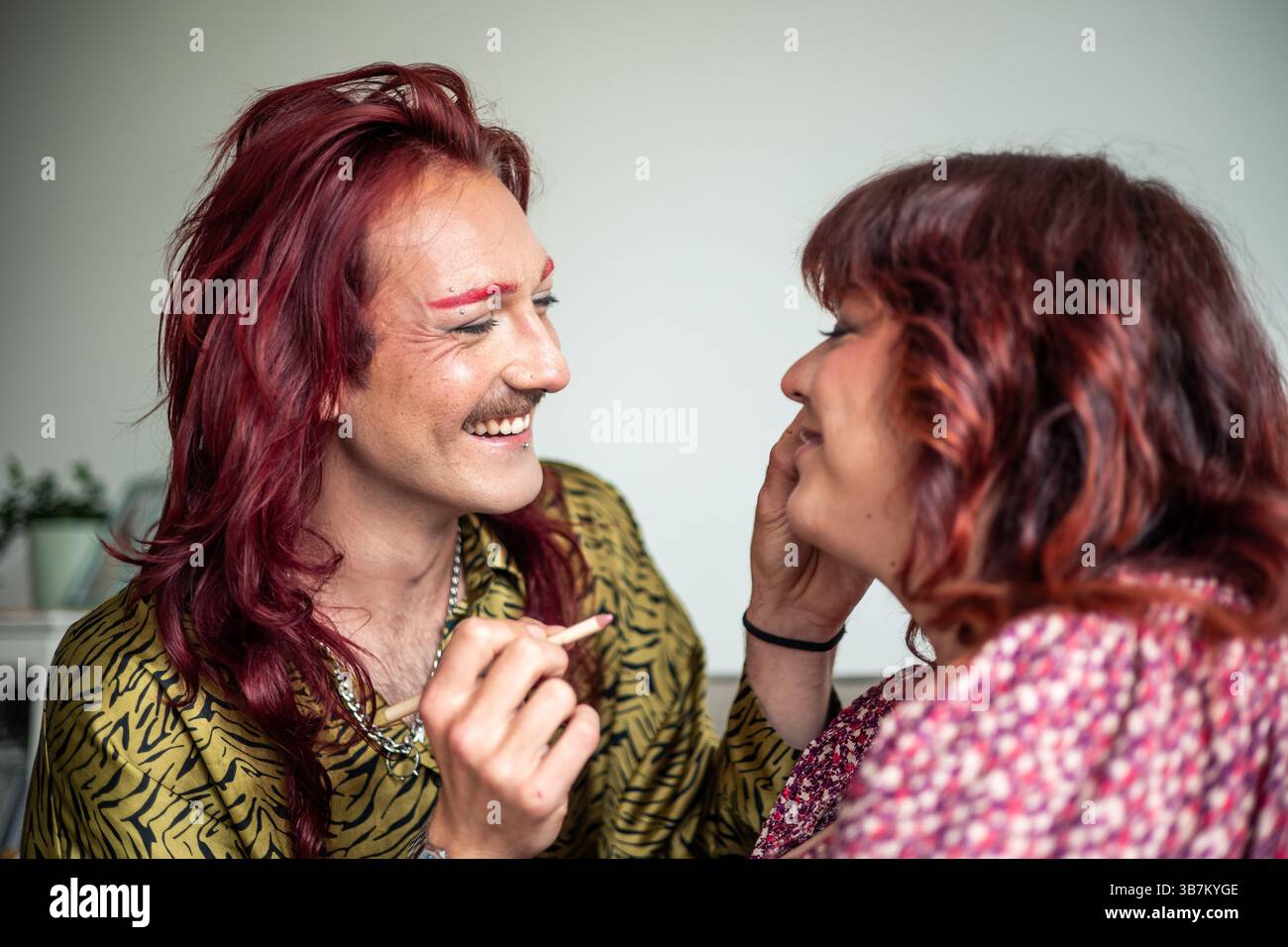 Smiling non-binary person with long red hair and makeup is applying makeup to their friend ...