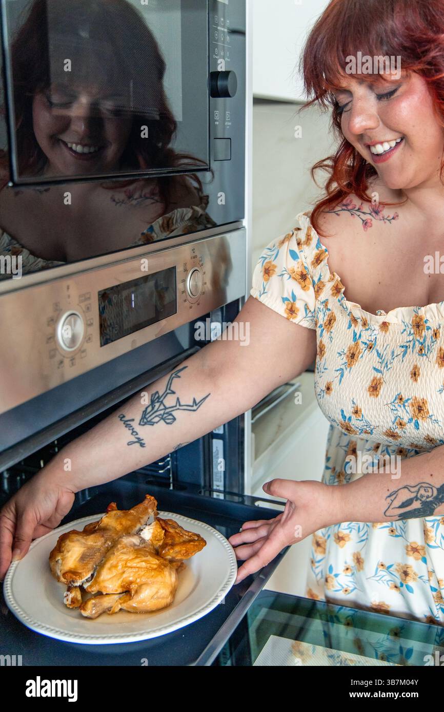 Happy young woman with red hair and a curvy figure taking a delicious roast chicken out of the ...