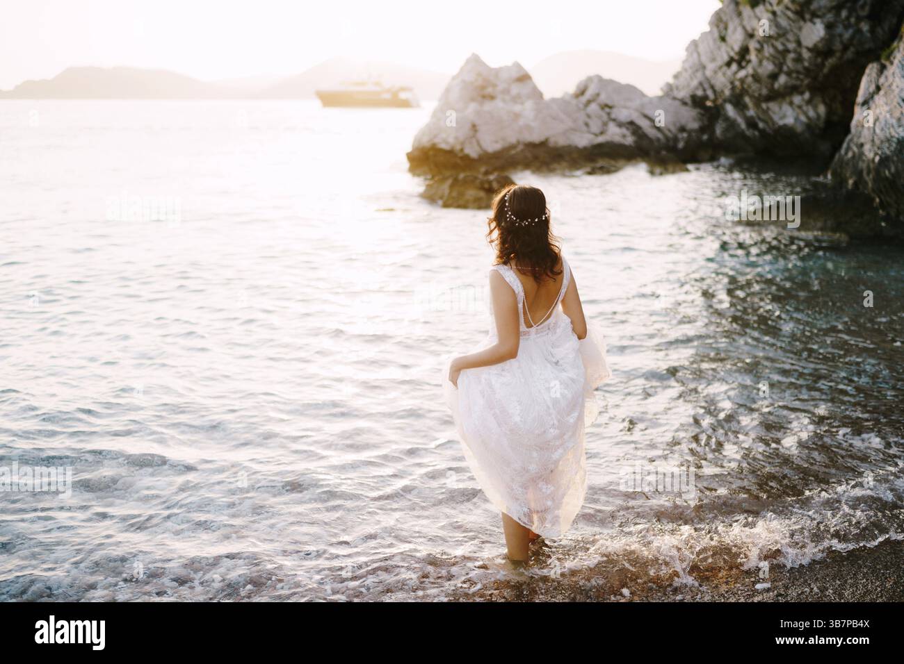 Bride stands in the sea, holding the hem of her white dress. Back view ...