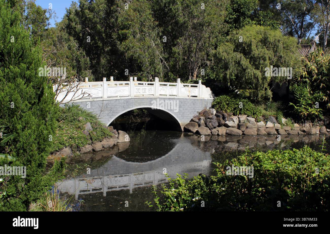 White arched bridge over a pond of water surrounded by plants and trees ...