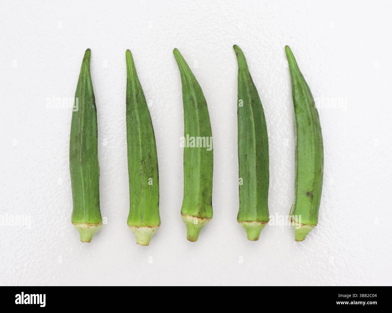 Five fresh, bright green okra pods are aligned in a row on a clean ...
