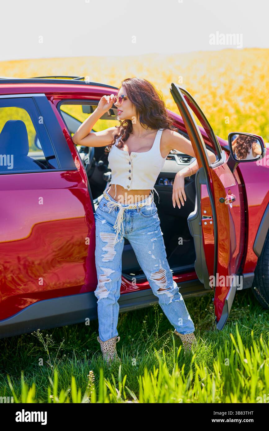 A beautiful woman with long hair stands next to a red SUV in a farm ...