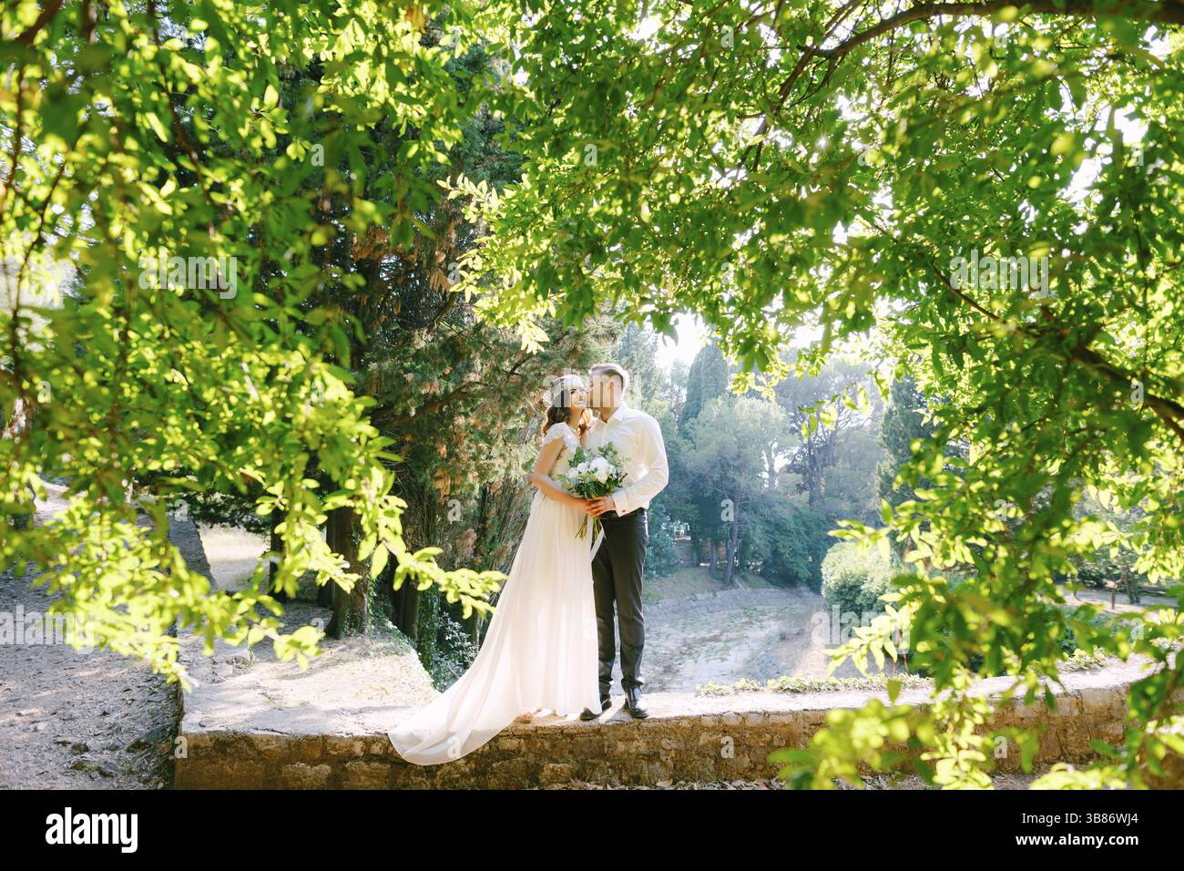 The bride and groom are hugging among beautiful lush branches of trees ...