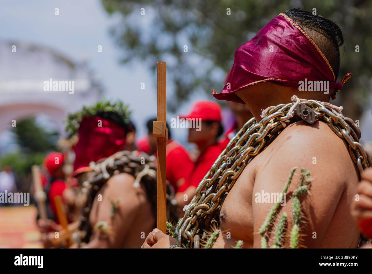 Man with covered face carries a wooden cross, wearing a crown of thorns and chains on his body ...