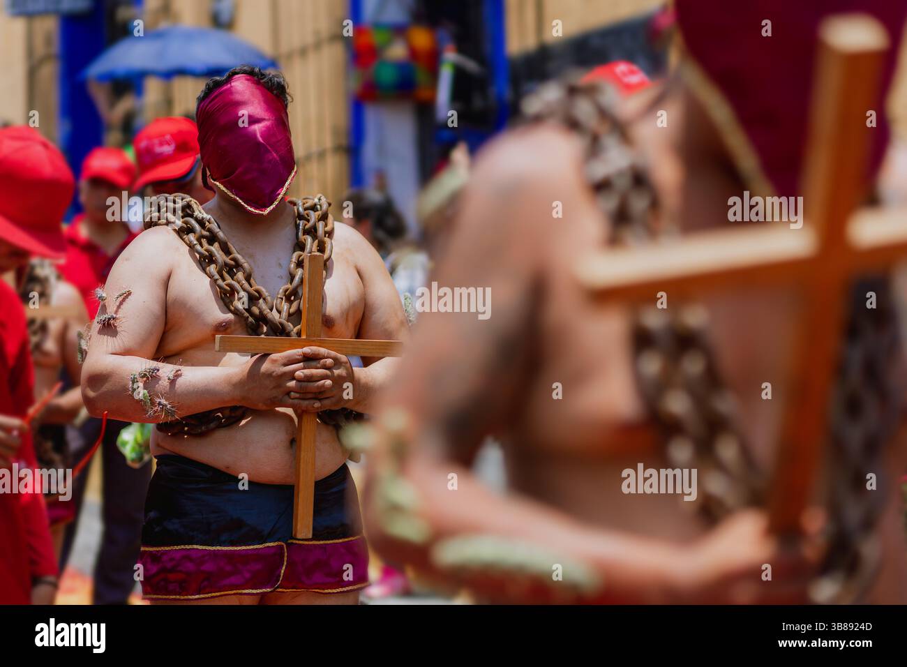 Man with covered face carries a wooden cross, wearing a crown of thorns and chains on his body ...