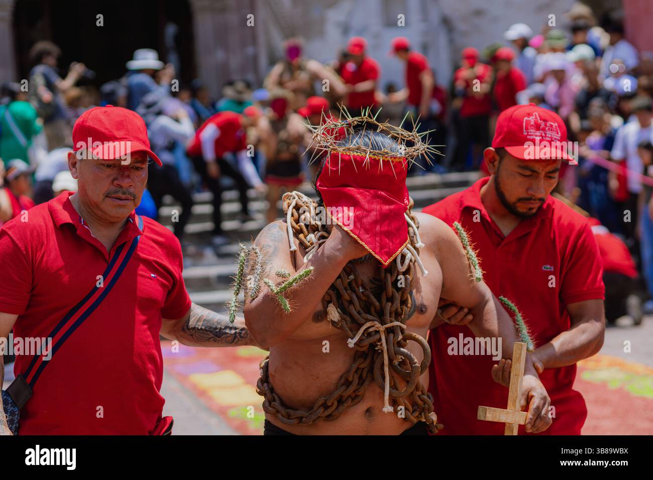 Man with covered face carries a wooden cross, wearing a crown of thorns and chains on his body ...
