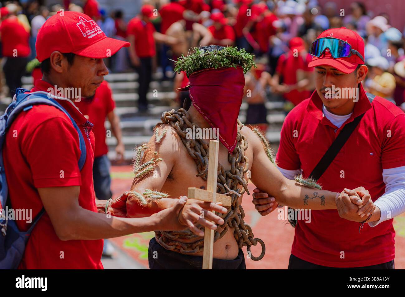Man with covered face carries a wooden cross, wearing a crown of thorns and chains on his body ...