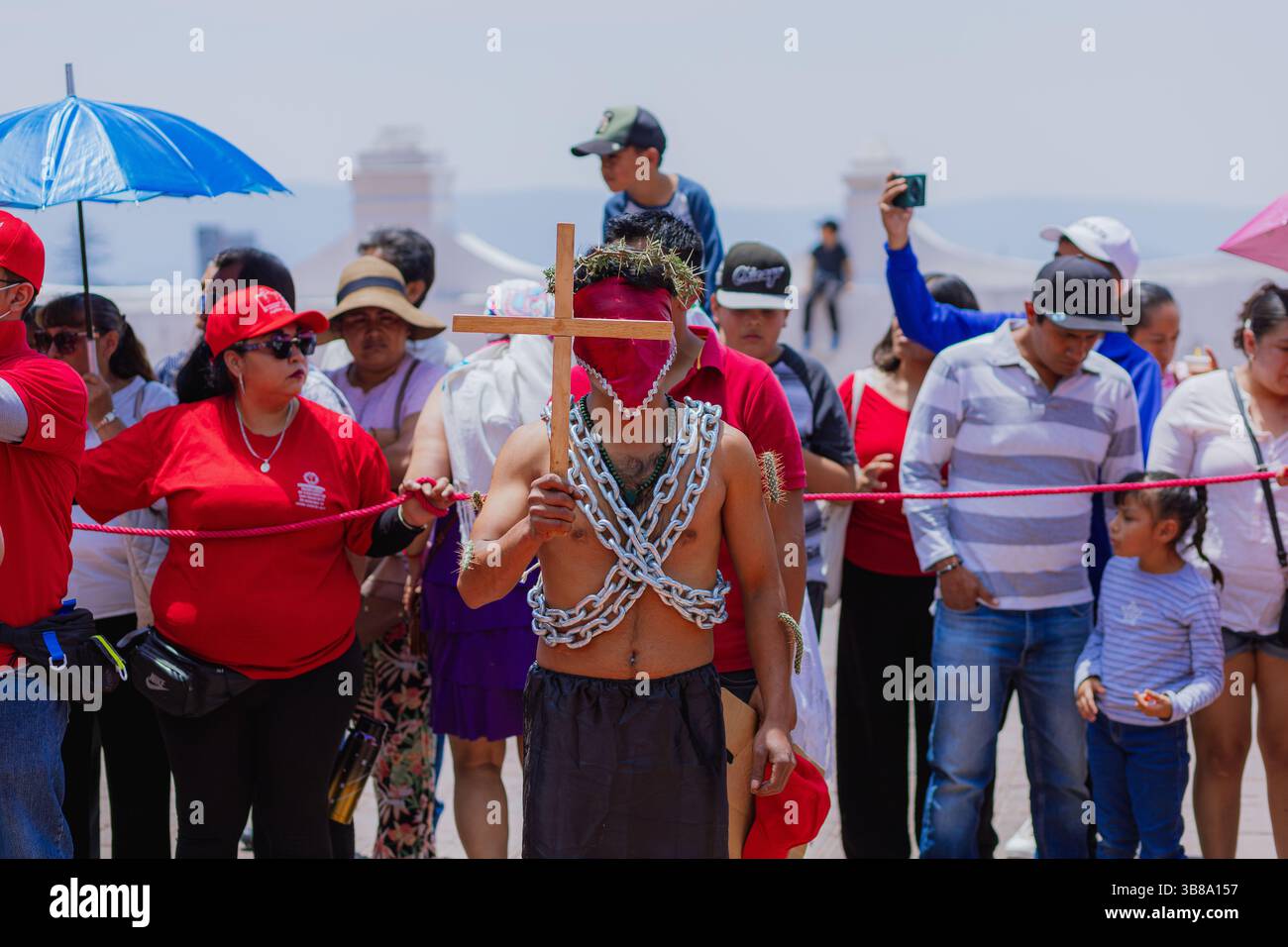 Man with covered face carries a wooden cross, wearing a crown of thorns and chains on his body ...