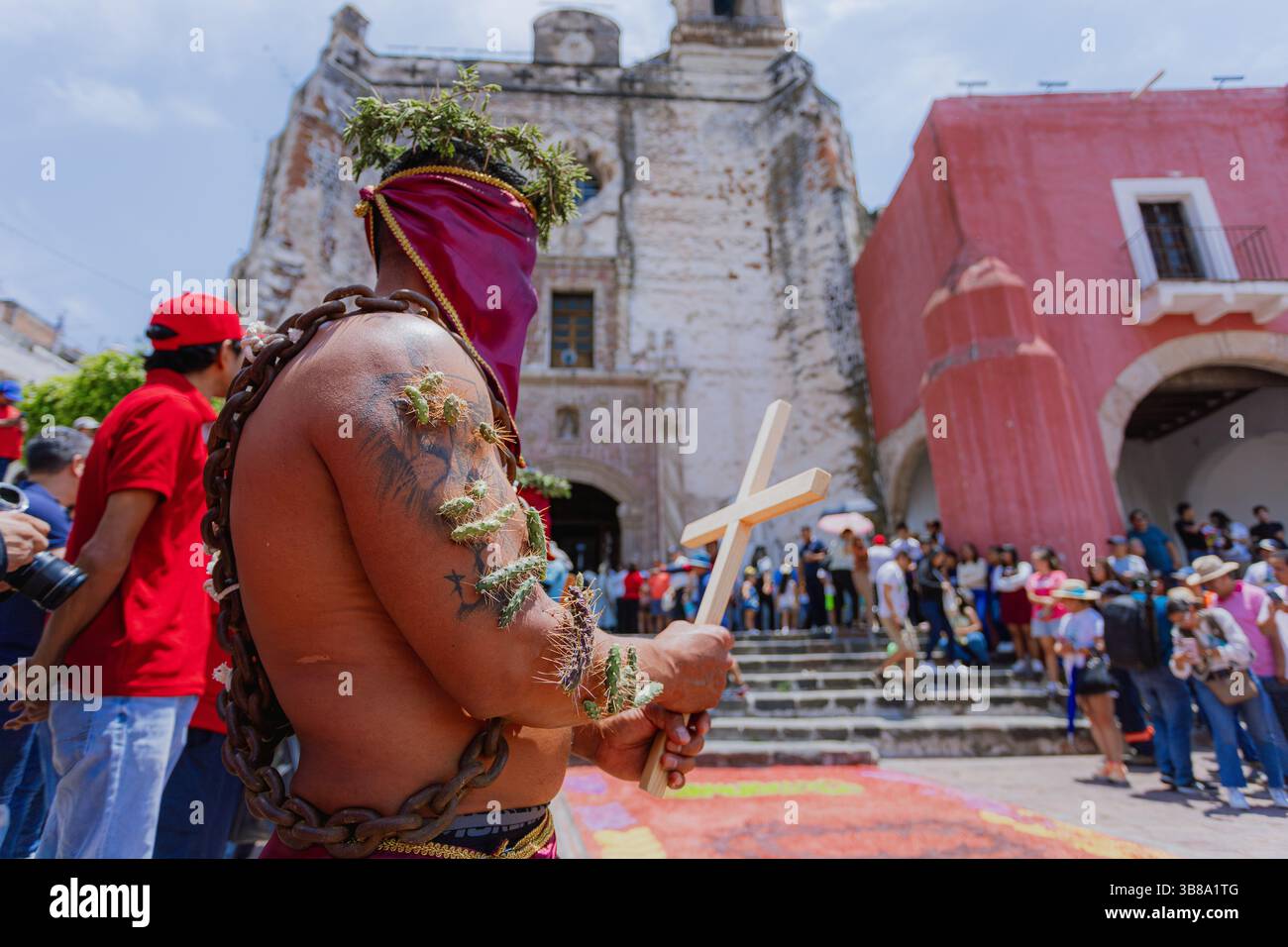 Man with covered face carries a wooden cross, wearing a crown of thorns and chains on his body ...