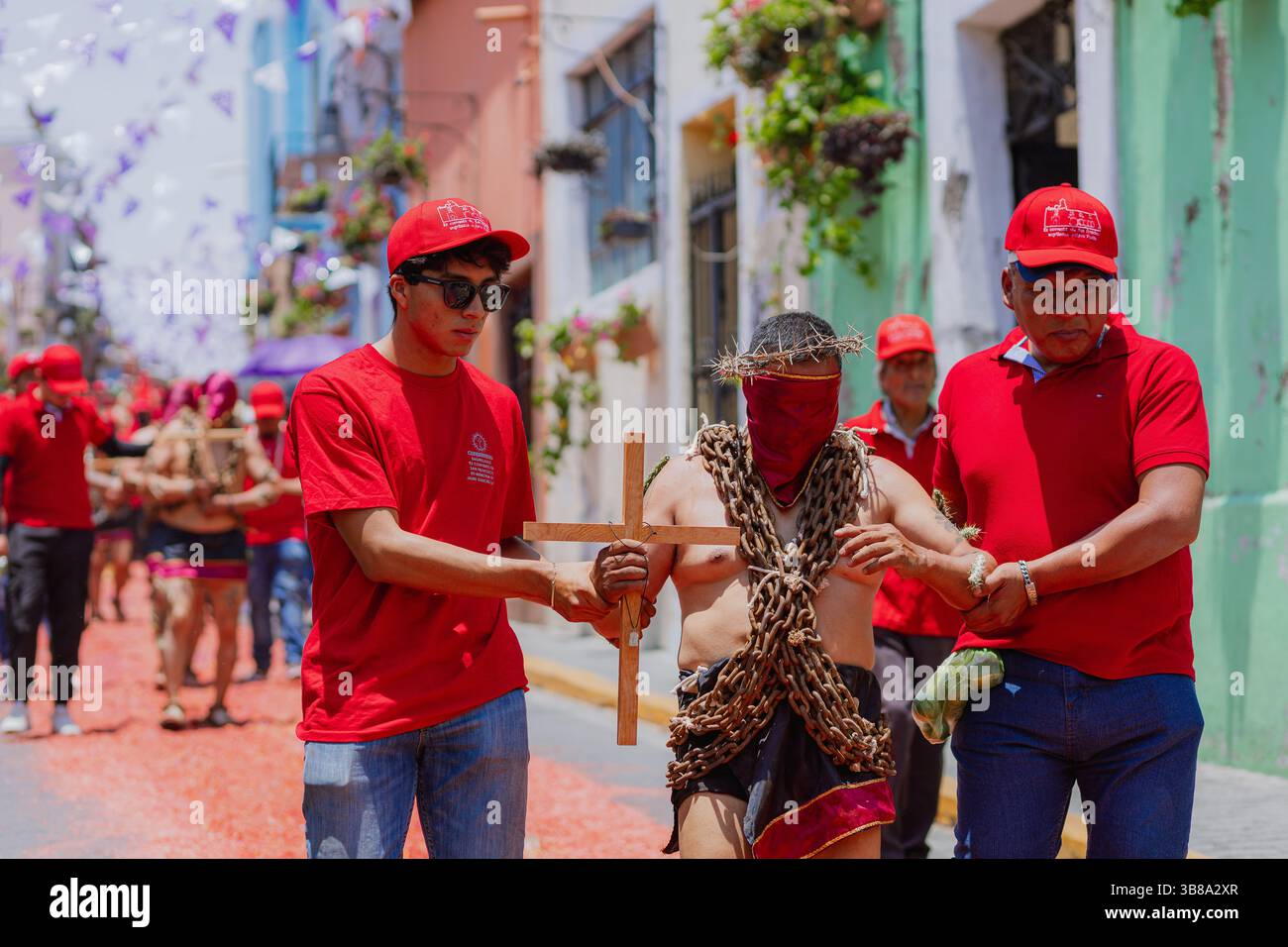 Man with covered face carries a wooden cross, wearing a crown of thorns and chains on his body ...