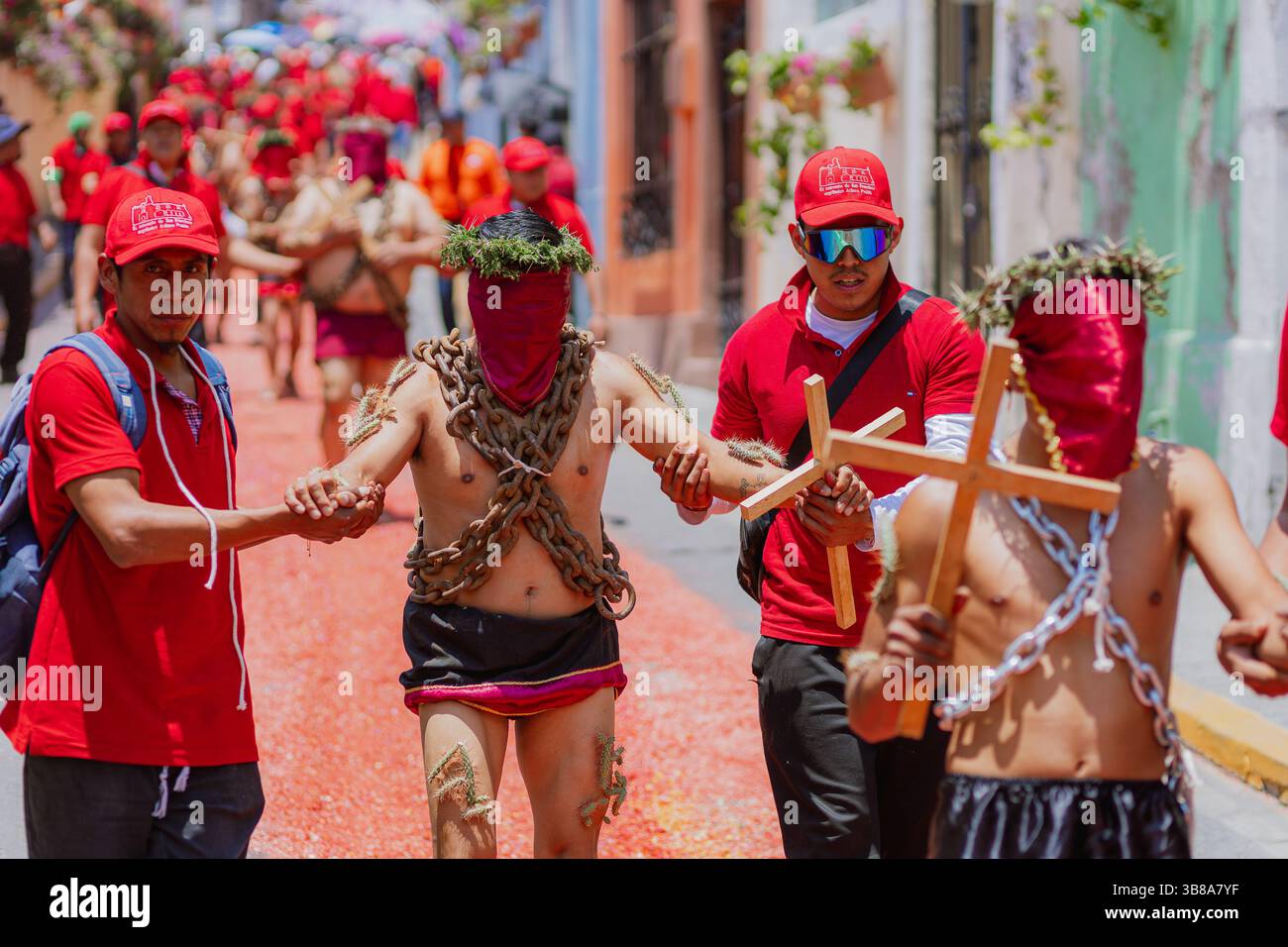 Man with covered face carries a wooden cross, wearing a crown of thorns and chains on his body ...