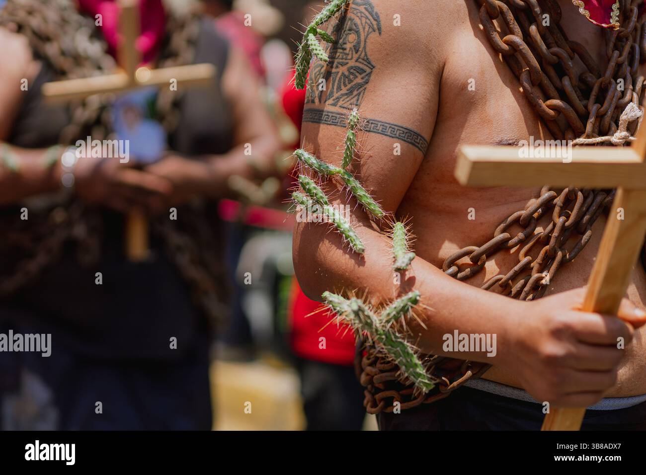 Man with covered face carries a wooden cross, wearing a crown of thorns and chains on his body ...