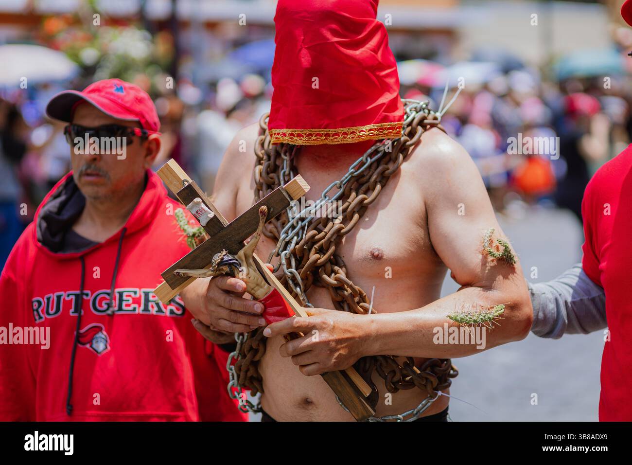 Man with covered face carries a wooden cross, wearing a crown of thorns and chains on his body ...