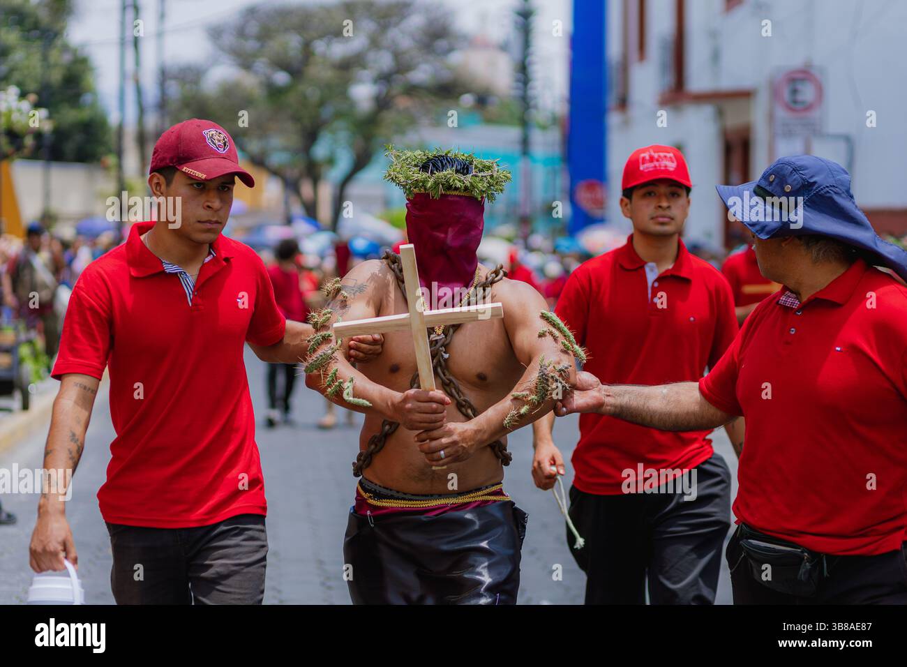 Man with covered face carries a wooden cross, wearing a crown of thorns and chains on his body ...