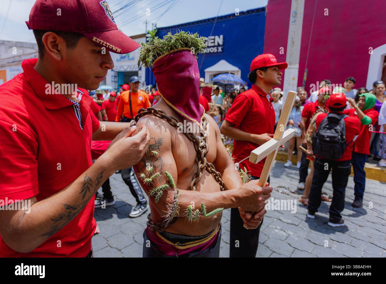 Man with covered face carries a wooden cross, wearing a crown of thorns and chains on his body ...