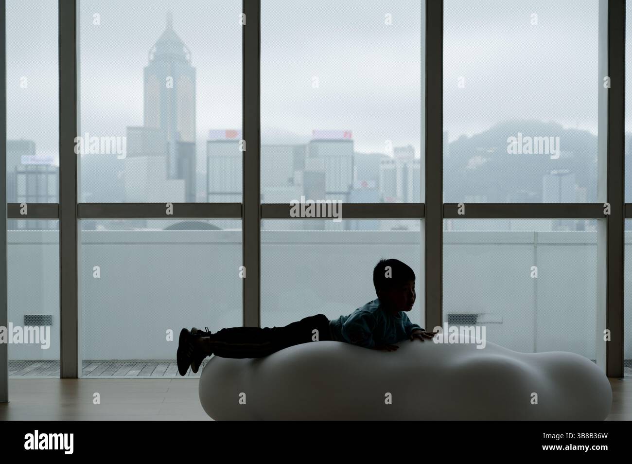 Kid lays on a cloud-shaped seat while looking out at the foggy Hong ...
